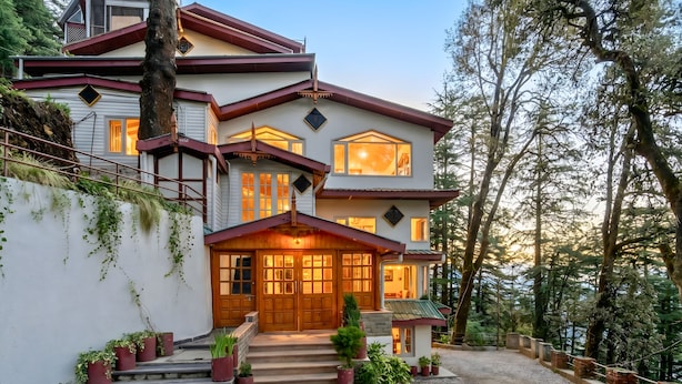Uma casa de dois andares com uma entrada frontal destacada, uma escadaria que leva à porta principal e uma paisagem ao redor com árvores.
