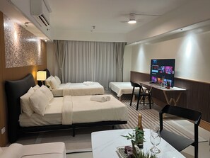 Quarto de hotel com duas camas, uma mesa de trabalho com uma TV, uma cadeira e uma pequena mesa com uma garrafa e copos.