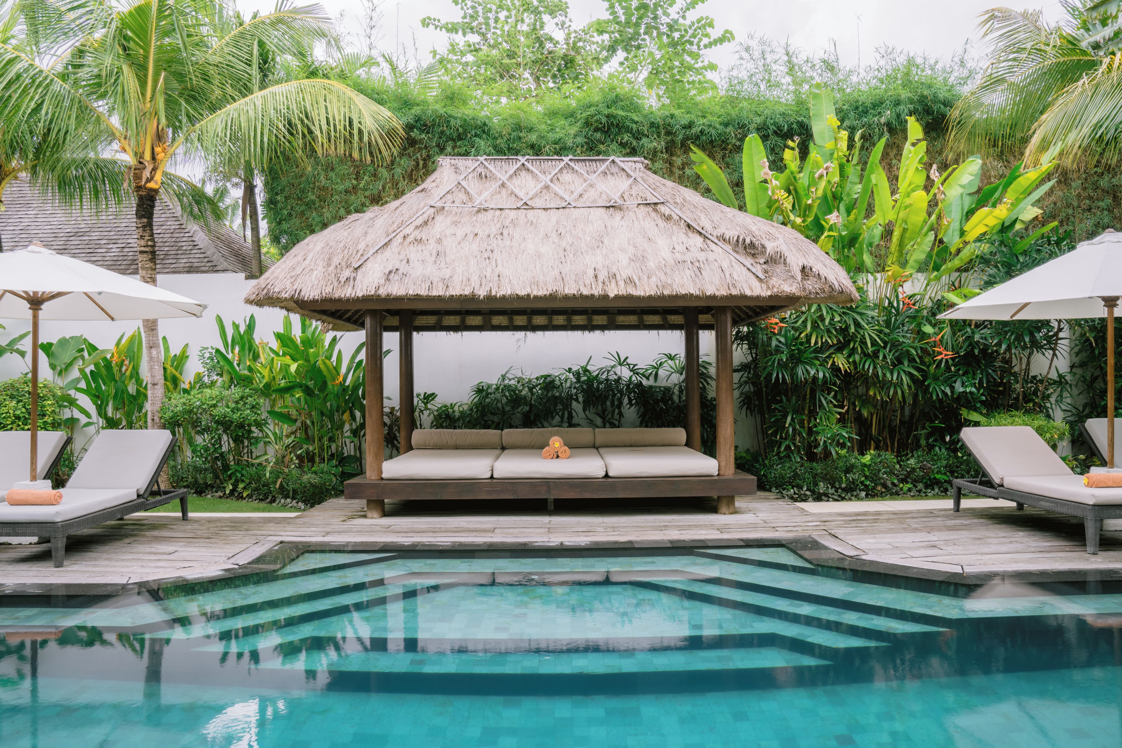 Uma área na beira da piscina com um bangalô de palha, espreguiçadeiras e piscina.