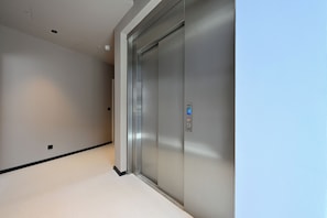 Um corredor com elevador moderno, piso claro e parede sem decoração.