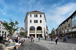 Um edifício com o nome 'WEMPE' na fachada, rodeado por uma praça de calçada e pedestres, e uma fonte.