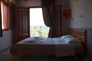 Uma cama de solteiro com cabeceira de madeira, porta de varanda e uma pequena mesa com uma cadeira do lado de fora.