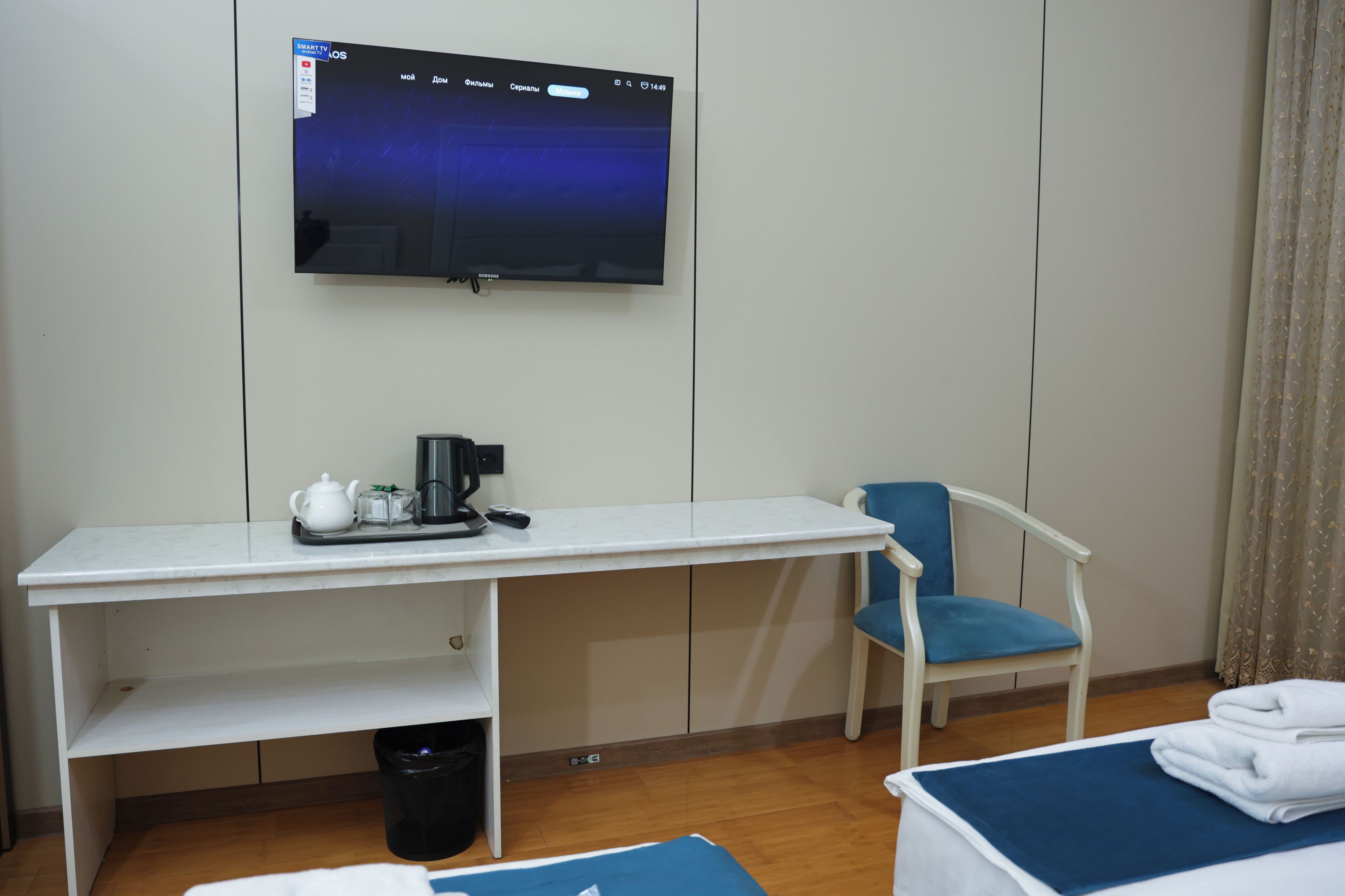 Quarto de hotel com televisão de tela plana na parede, escrivaninha com cafeteira, cadeira e cama com manta azul.