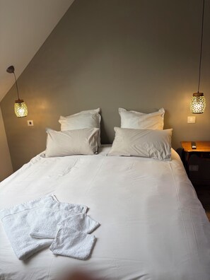 Cama bem arrumada com lençóis e almofadas brancos, duas luminárias pendentes com cúpulas estampadas e uma mesa de cabeceira de madeira com um controle remoto.