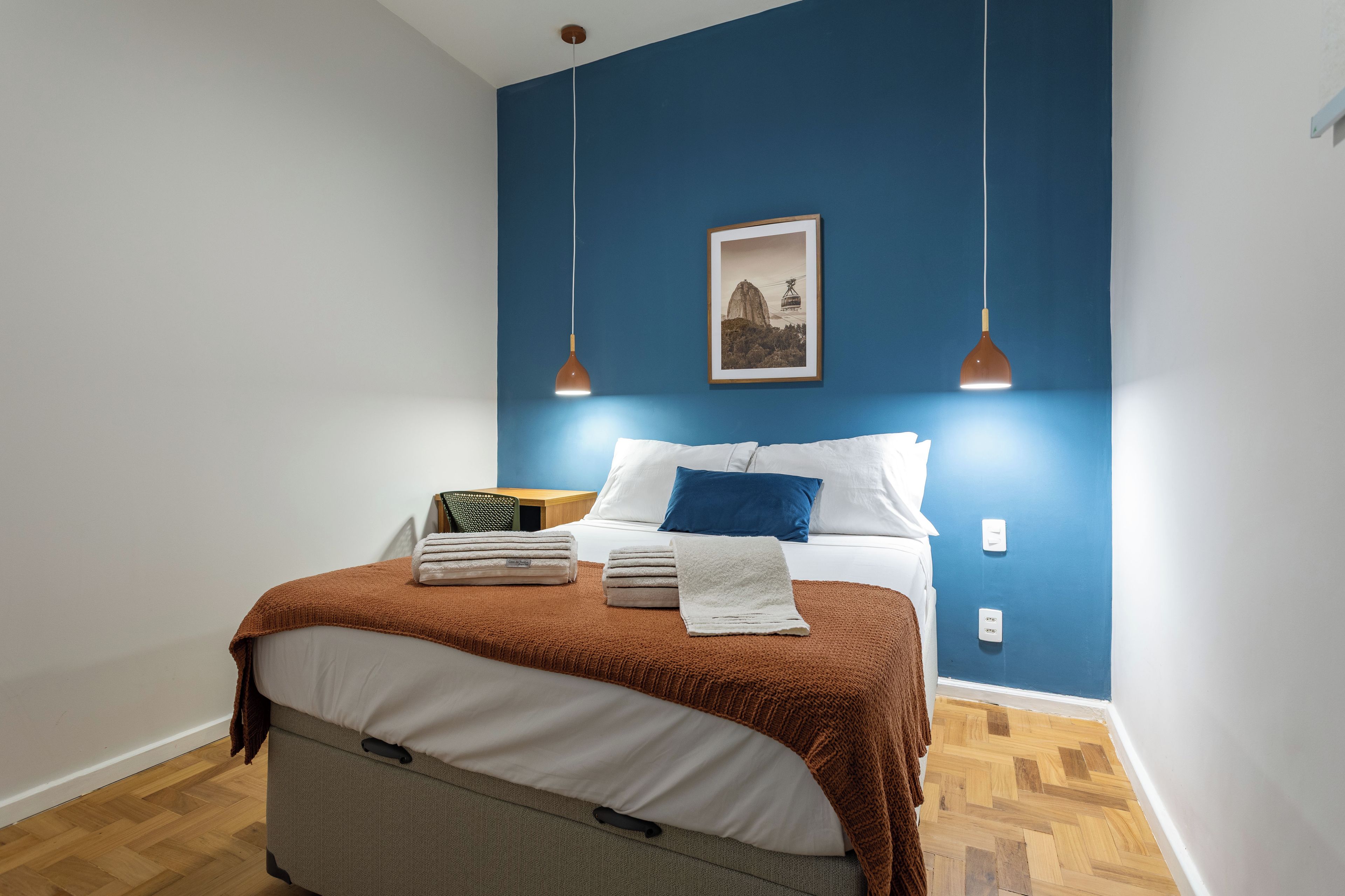 Cama com cobertor castanho, almofadas brancas e uma almofada azul. Há um quadro na parede acima da cama.