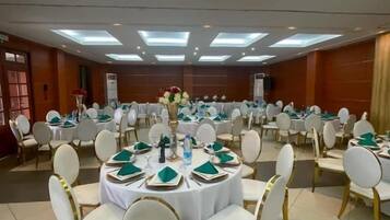 Uma sala de banquetes com mesas redondas, cadeiras brancas e guardanapos verdes.