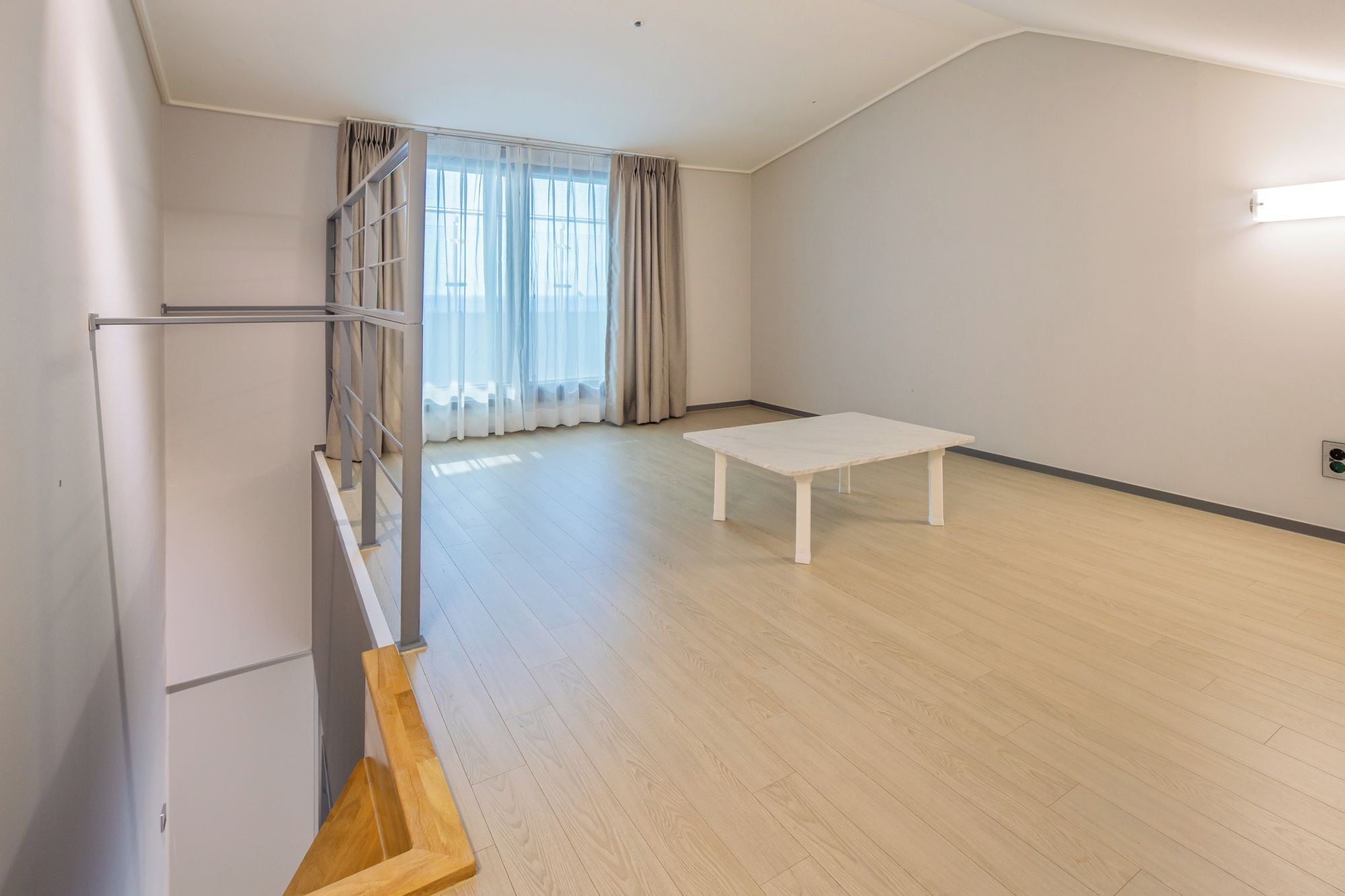 Um quarto espaçoso com piso de madeira, uma mesa branca e uma janela com cortinas.