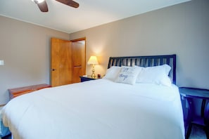 Quarto de hotel com uma cama grande, mesa de cabeceira com abajur, porta de madeira e ventoinha no teto.