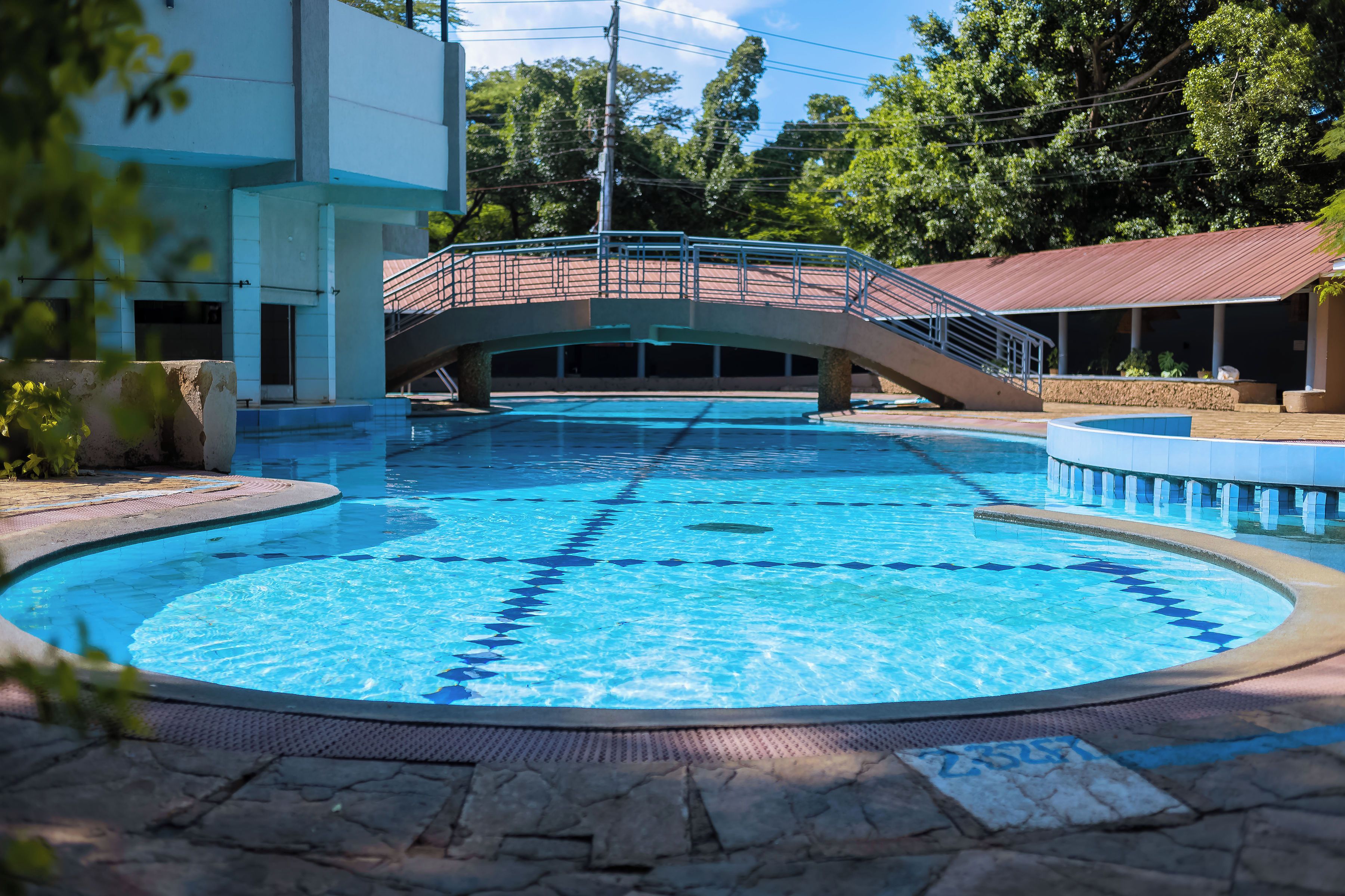 Piscina com uma ponte, rodeada por árvores e um edifício.