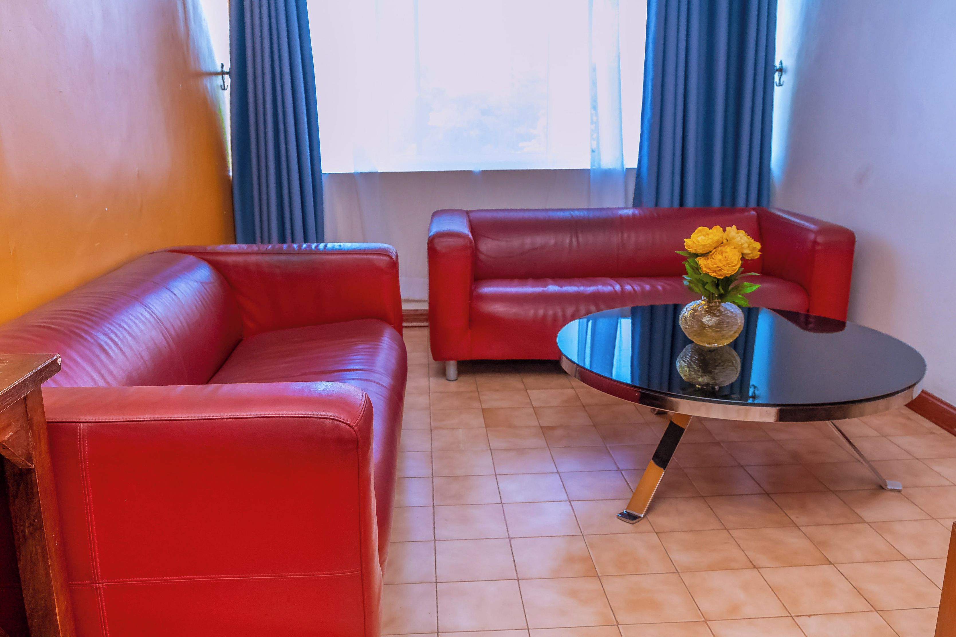 Um quarto com dois sofás de couro vermelho, uma mesa de centro de vidro com um vaso de flores amarelas e uma janela com cortinas azuis.