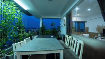 Espaço de refeições com mesa, cadeiras, um ventilador e vista para a cidade ao entardecer.