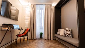 Quarto de hotel moderno com televisão de tela plana, escrivaninha com laptop, cadeira vermelha, sofá com almofadas estampadas e planta em vaso.