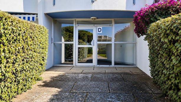 Entrada de um prédio com porta de vidro identificada pela letra 'D', cercada por vegetação e céu azul límpido.