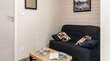 Quarto com sofá preto, duas almofadas estampadas, mesa de centro de madeira com revista e livro, quadro na parede e porta com puxador.