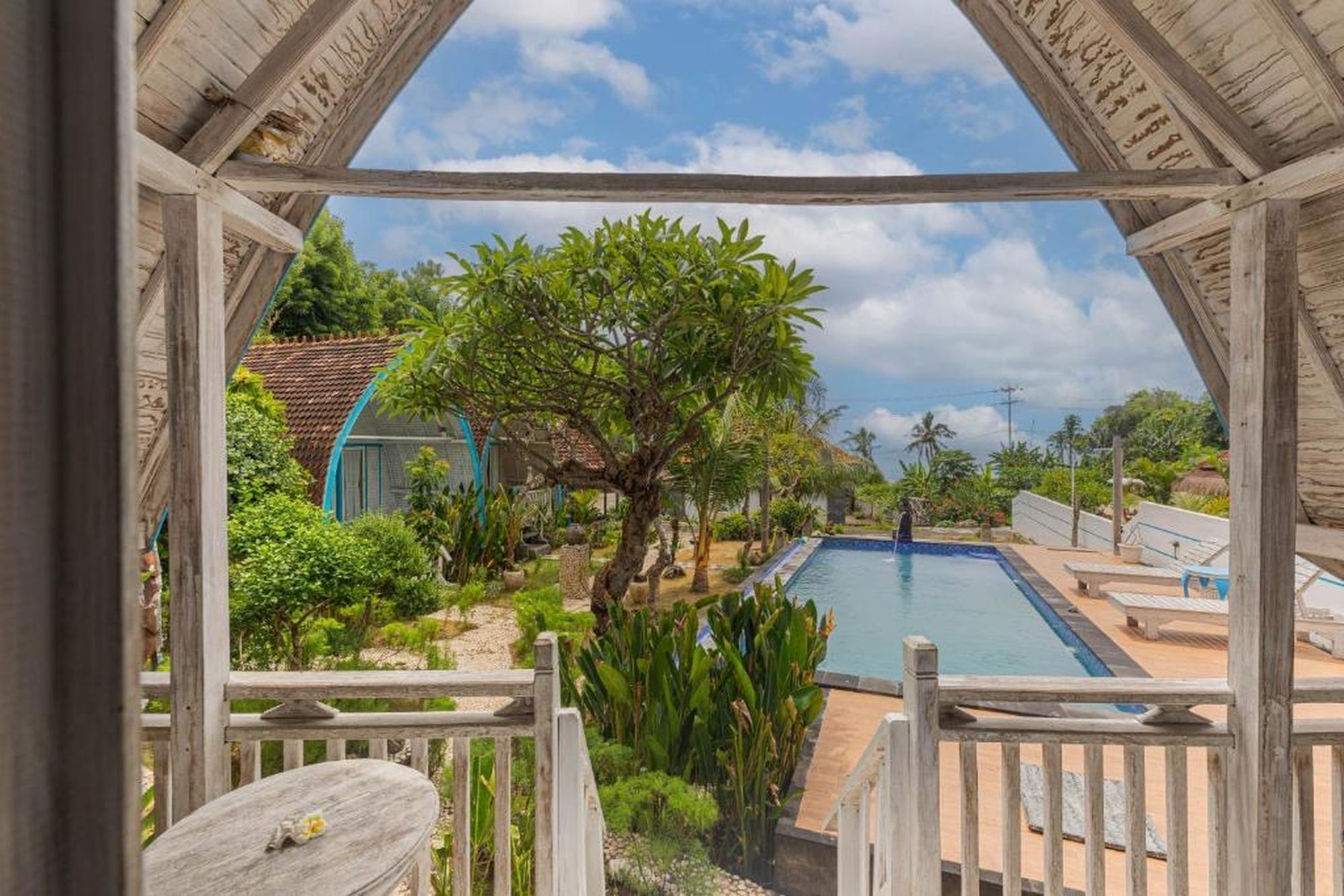 Uma área de piscina com deck de madeira, espreguiçadeiras e vista para um jardim com árvores e um edifício ao fundo.