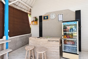 Uma área de recepção com balcão de madeira, dois banquinhos, uma geladeira repleta de bebidas e um relógio na parede.