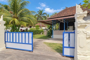 Uma entrada protegida por grades, com varanda coberta, palmeiras e céu azul.