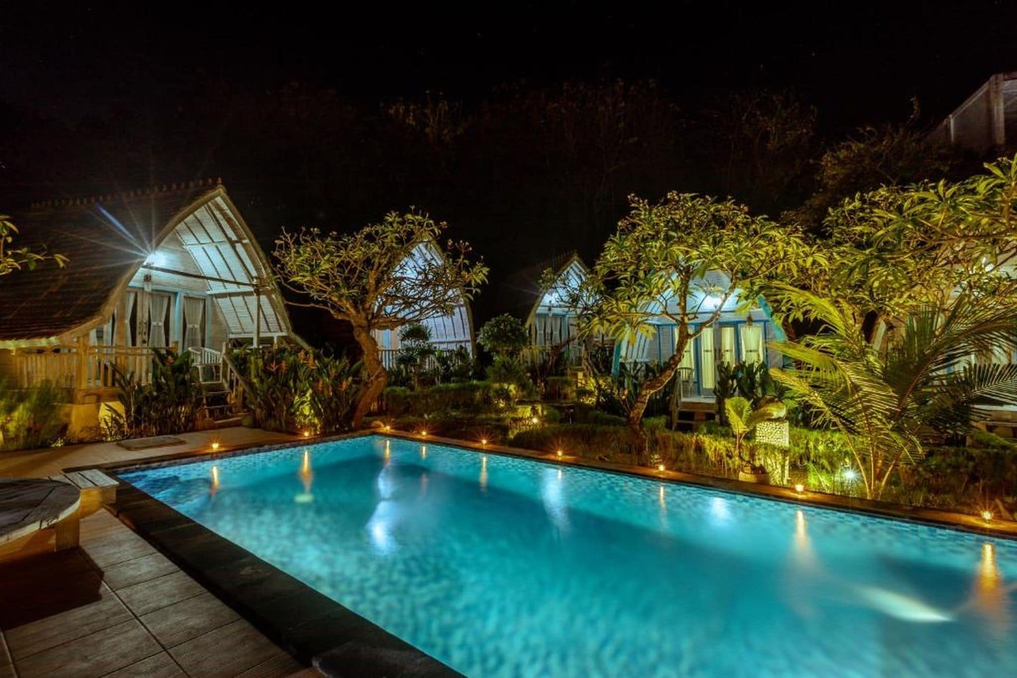 Área de piscina à noite, com um edifício de cobertura de vidro e vegetação tropical.