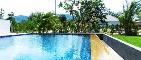 Piscina com água azul cristalina, cercada por vegetação e um edifício ao fundo.