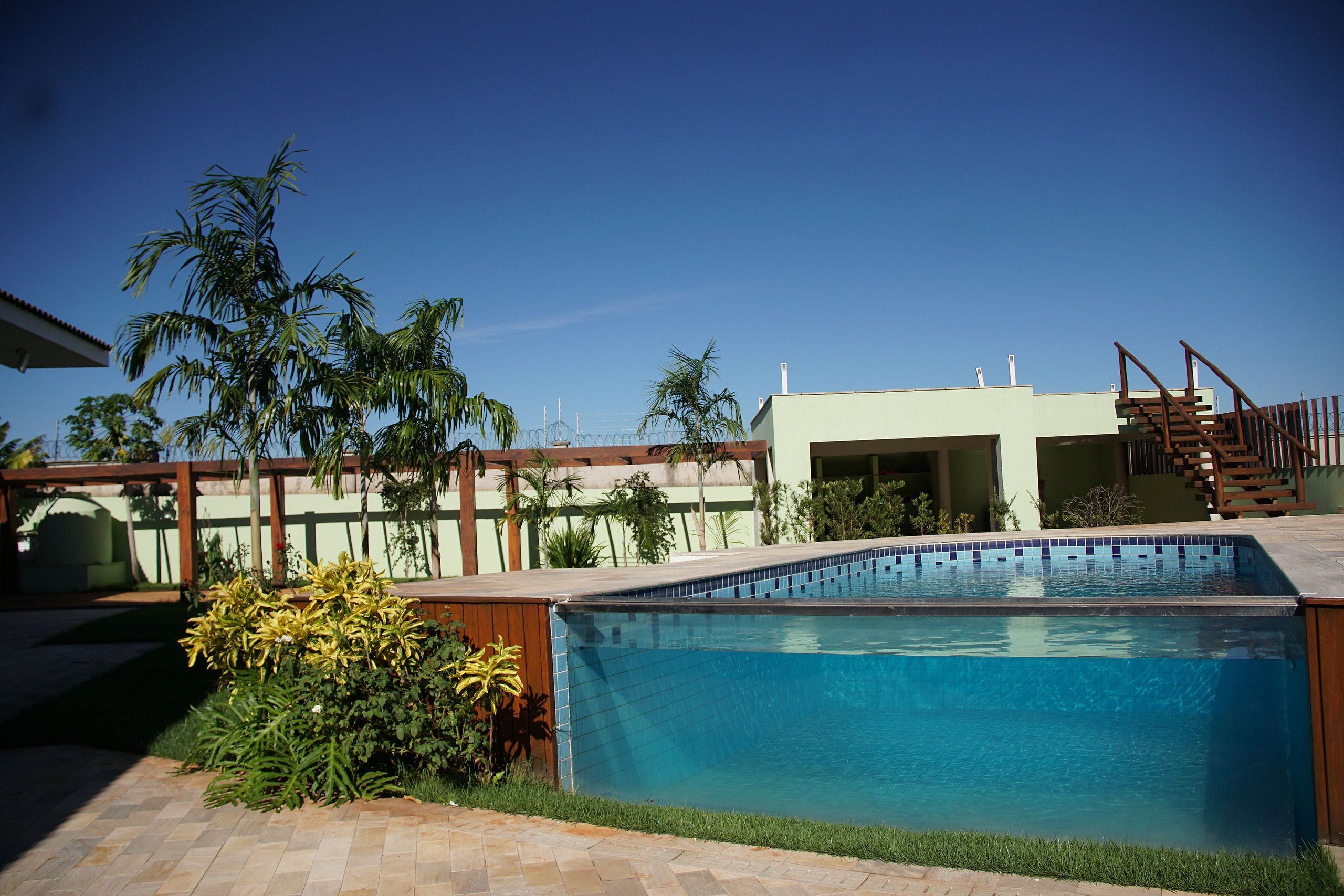 Uma piscina com borda azulejada, rodeada por vegetação e um edifício com escadas.