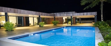 Uma área de piscina moderna com piscina azul cristalina, rodeada por um deck de madeira e portas de vidro que dão para um edifício.
