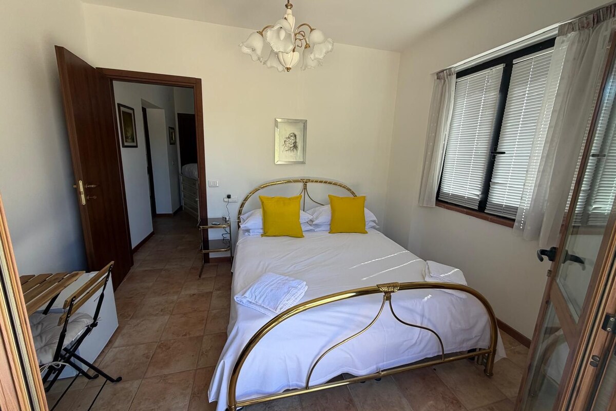 Quarto com cama, mesa de cabeceira, abajur e janela com persianas.