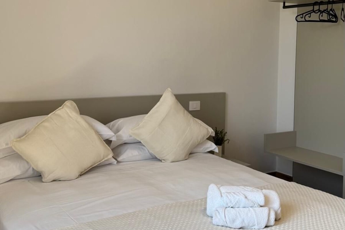 Cama bem arrumada com almofadas e uma toalha dobrada, mesa de cabeceira e prateleira na parede.