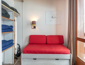 Quarto compacto com cama, prateleira com roupas dobradas, luminária na parede e quadro emoldurado.