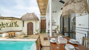 Área da piscina com uma mesa de jantar posta para dois, uma estrutura com telhado de palha e um edifício branco com porta de madeira.