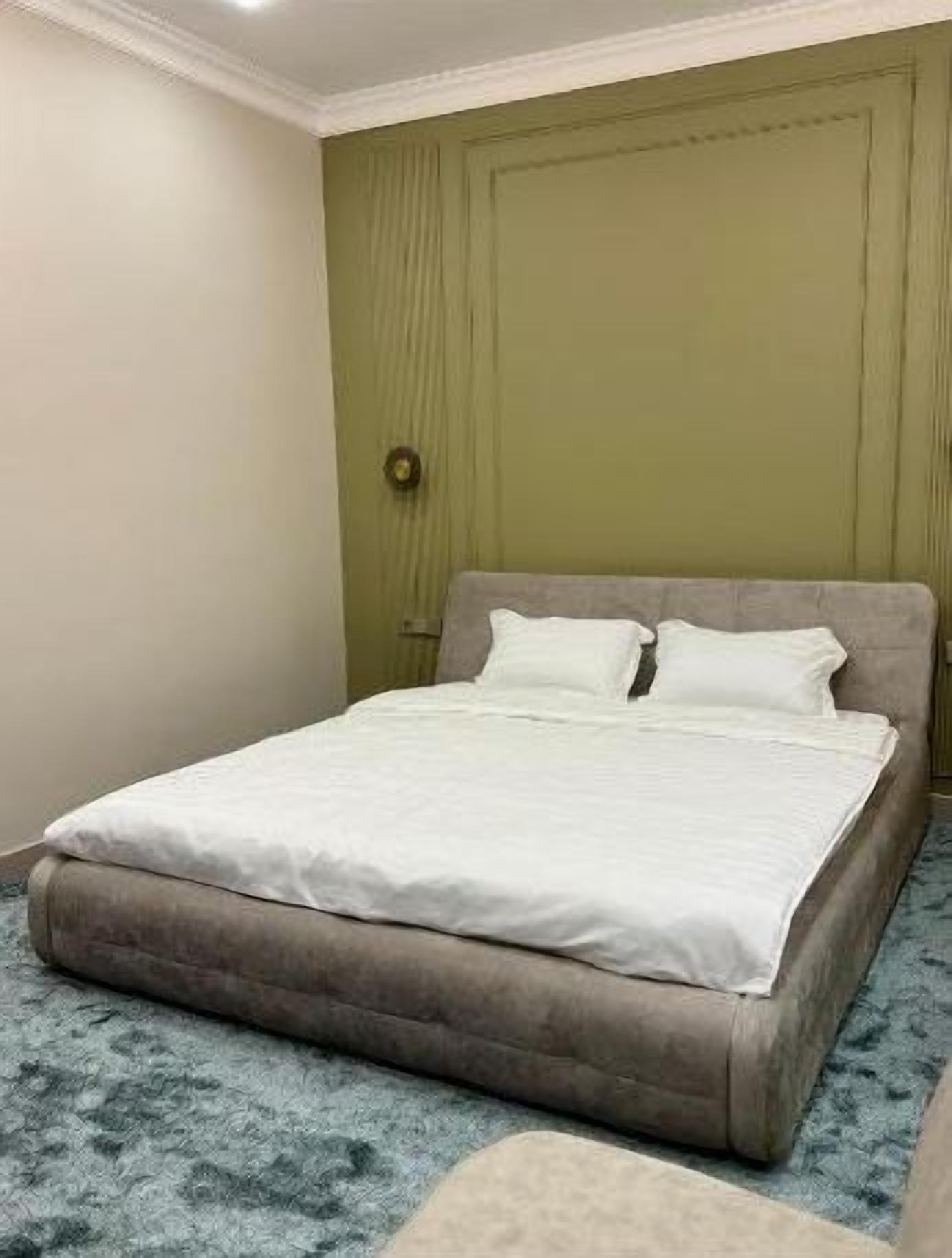 Cama com colcha branca e duas almofadas, porta com puxador e chão coberto por carpete.