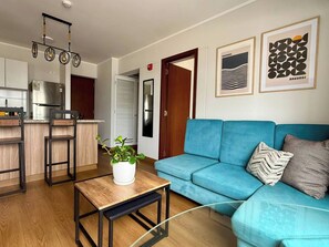 Sala de estar moderna com sofá azul, mesa de centro e área de cozinha ao fundo.