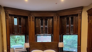 Quarto com cama grande, duas mesas de cabeceira, radiador, janela com persianas e vista para área verde, além de um tapete estampado.