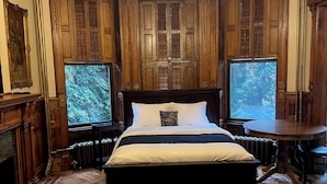 Quarto com cama grande, duas mesas de cabeceira, um lustre e vista para uma floresta pela janela.