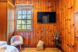 Quarto com paredes de madeira, uma cama, uma cadeira, uma mesinha com um vaso e uma televisão fixada na parede.