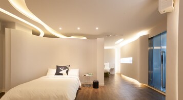 Quarto moderno com cama, mesa de cabeceira, lixeira e aquecedor fixado na parede.
