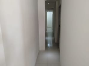 Um corredor estreito, com piso de azulejos, paredes brancas e uma porta no final.