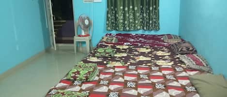 Quarto com cama coberta por roupa de cama estampada, um ventilador, uma janela com cortinas e uma porta que dá para outro quarto.