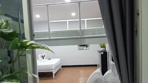 Quarto com cama, uma mesinha, planta em vaso e janela com cortinas.