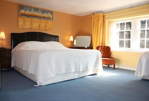 Quarto de hotel com uma cama grande, uma cadeira, escrivaninha com abajur e janela com cortinas.