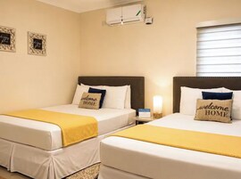 Quarto de hotel com duas camas, cada uma com almofada de 'welcome home', mesa de cabeceira com luminária e ar-condicionado fixado na parede.