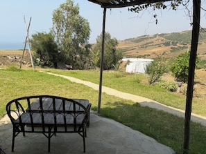 Uma área de estar coberta ao ar livre com um banco, um caminho de concreto e vista para uma paisagem montanhosa com vegetação e um edifício ao longe.