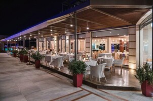 Uma área de refeições ao ar livre com mesas e cadeiras, uma galeria coberta e a entrada de um edifício.