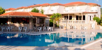 Piscina com espreguiçadeiras e área de bar, cercada por um edifício com telhado de telhas vermelhas.