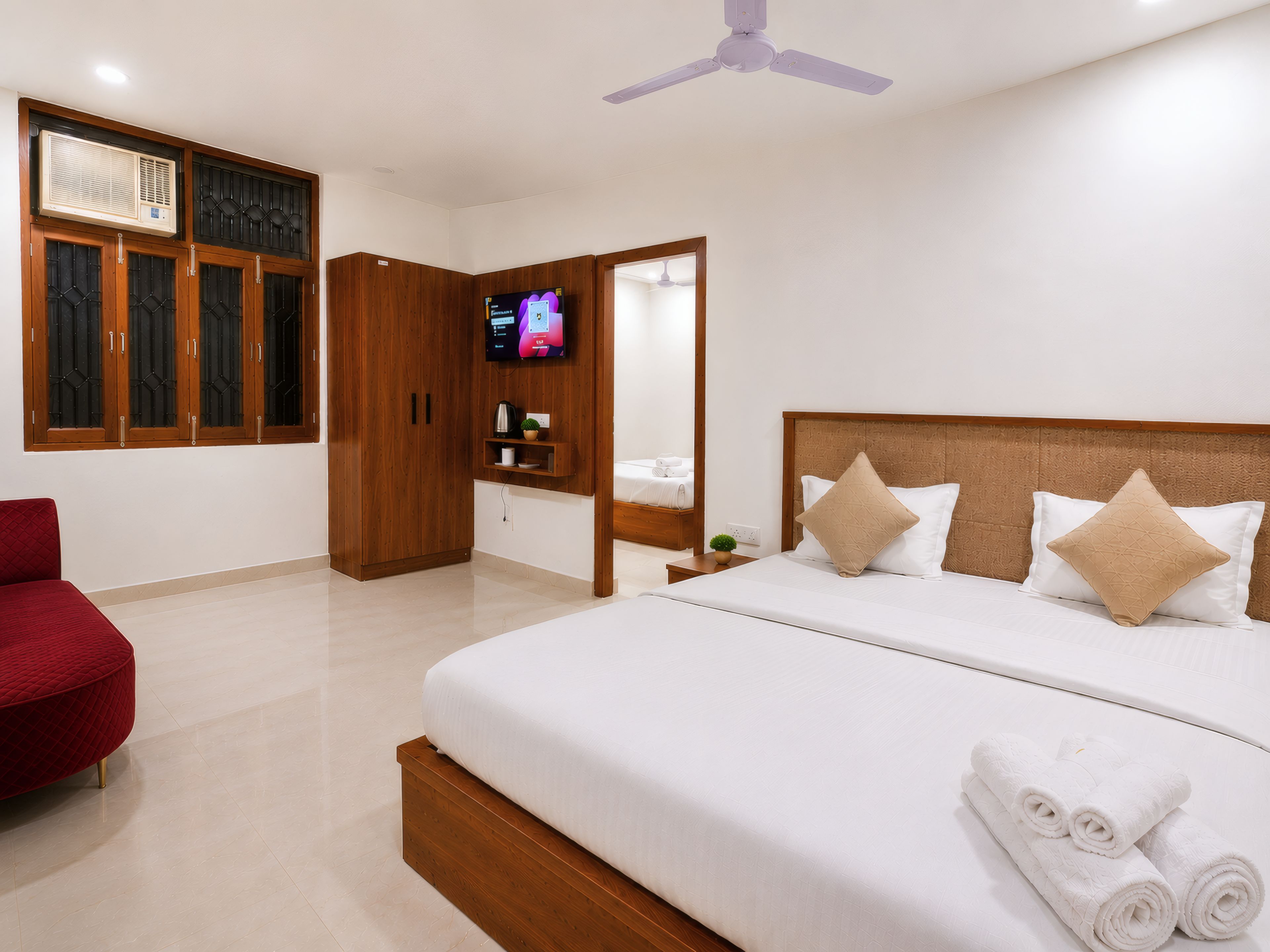 Quarto de hotel com cama, uma cadeira vermelha, guarda-roupa, televisão e ventoinha no teto.