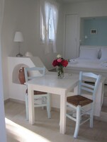 Um quarto com cama, uma mesa com cadeiras, um vaso com flores, um abajur e um quadro na parede.