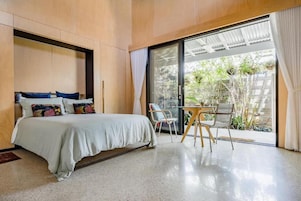 Quarto com cama, mesa e cadeiras, e porta de vidro deslizante para uma varanda com plantas.