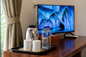 Mesa de madeira com uma bandeja contendo uma chaleira, duas xícaras e duas garrafas de água. Uma televisão exibindo uma imagem azul abstrata está fixada na parede atrás da mesa.