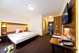 Quarto de hotel com uma cama, escrivaninha, cadeira, televisão e banheiro visível por uma porta aberta.