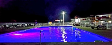 Piscina à noite com grade, espreguiçadeiras e um prédio ao fundo.