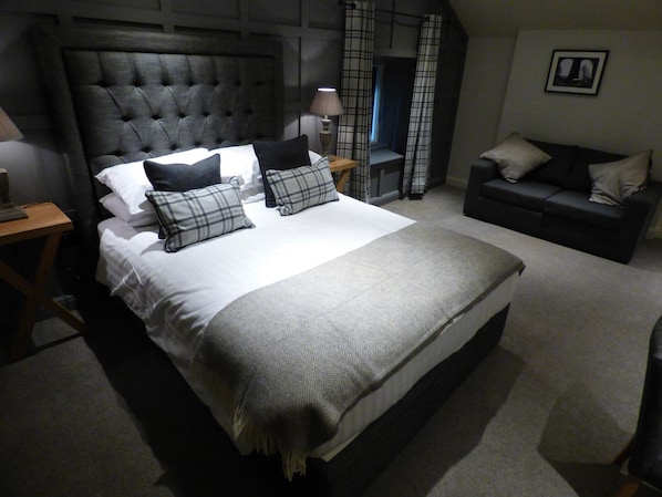 Quarto com uma cama grande, mesa de cabeceira, abajur, janela com cortinas, sofá e um quadro na parede.